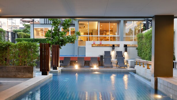 Piscina interior moderna com água azul cristalina, rodeada por espreguiçadeiras e um restaurante ao fundo.