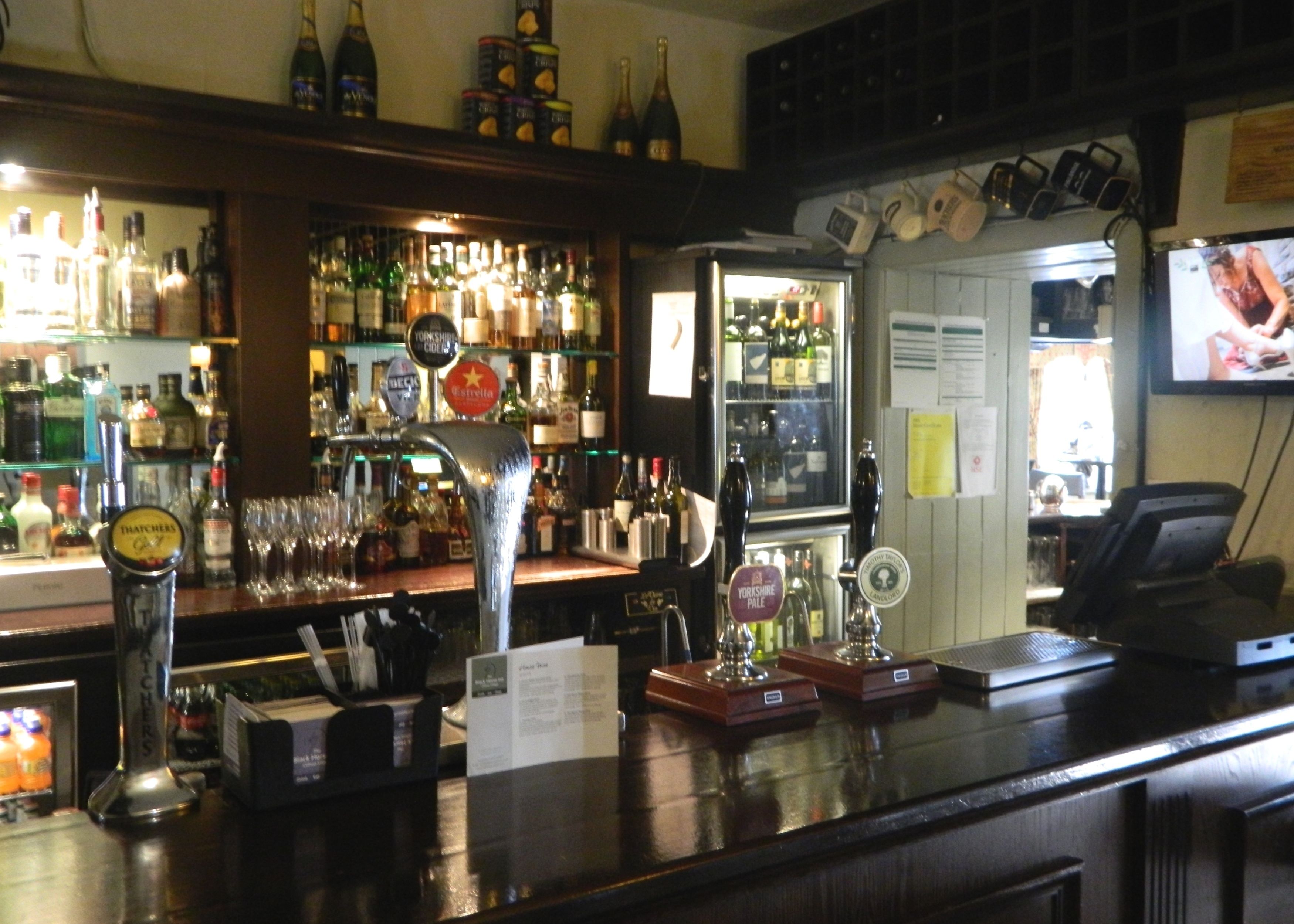 Uma área de bar com diversos frascos e copos, uma geladeira e uma televisão exibindo um programa.
