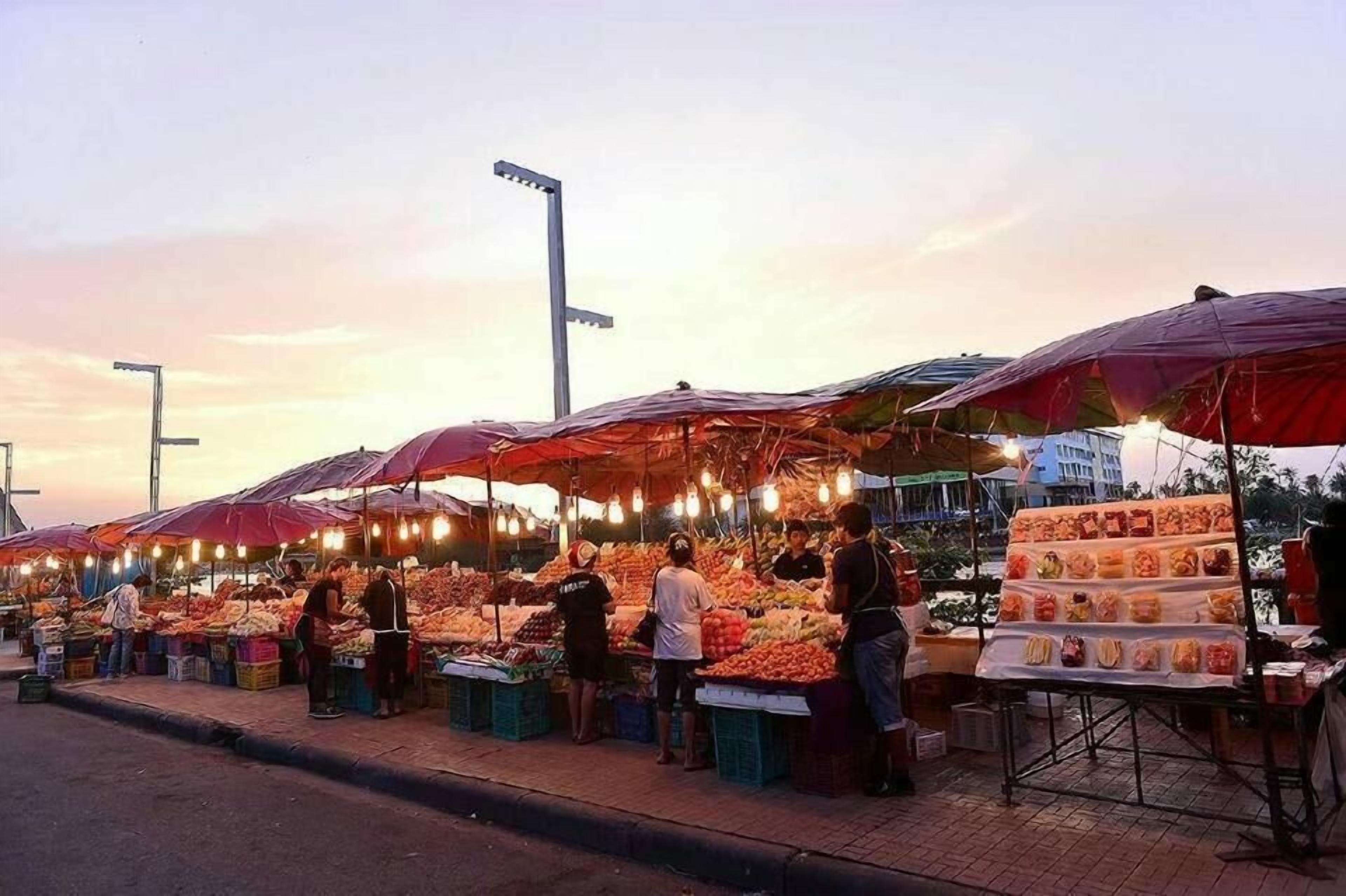 Mercado com bancas vendendo frutas e legumes sob guarda-sóis vermelhos.