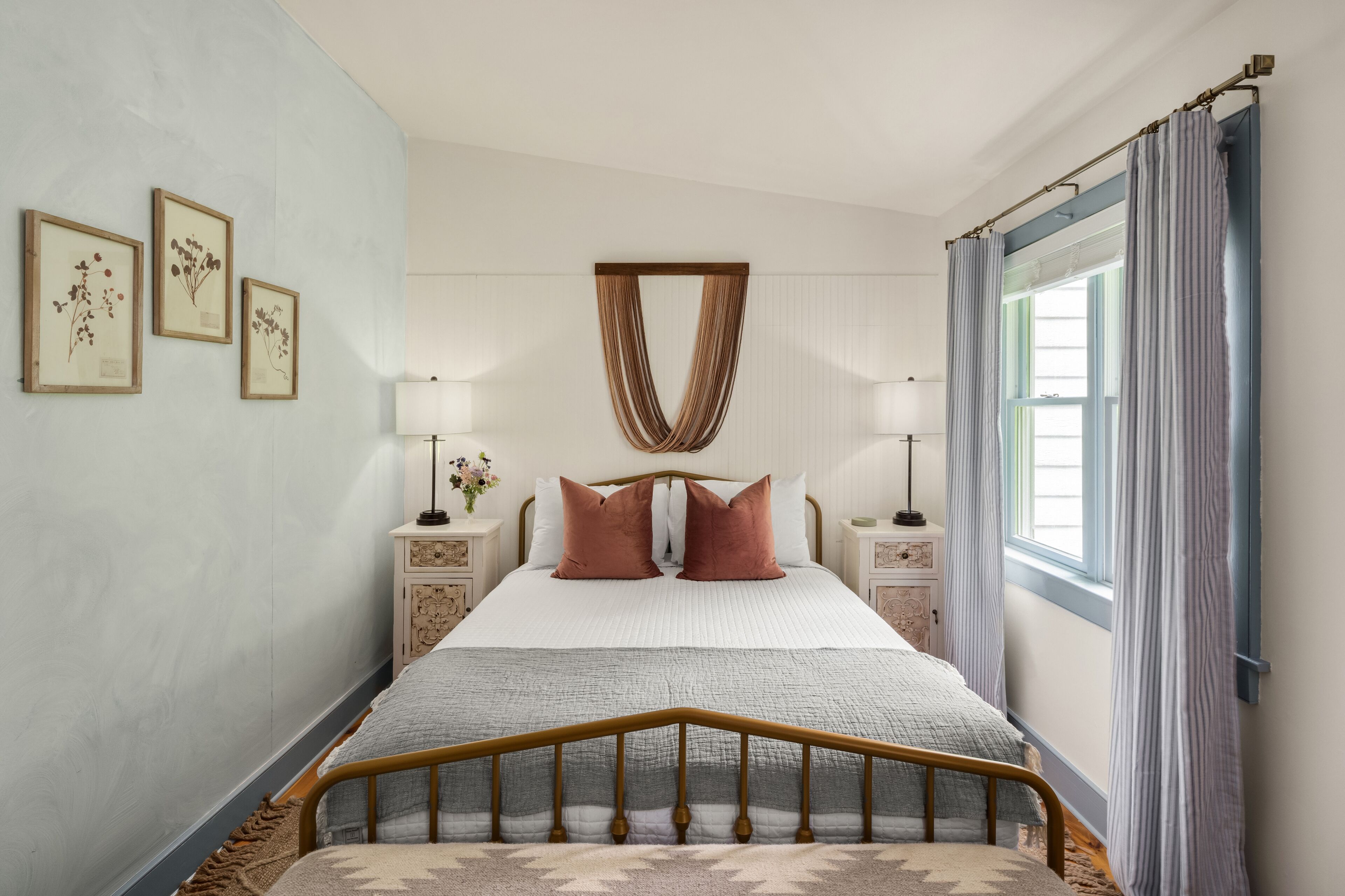 Quarto com cama, duas mesas de cabeceira, janela com cortinas e obra de arte na parede.