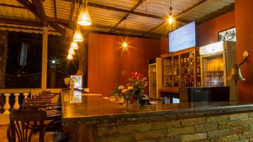 Uma área de bar com cadeiras de madeira, balcão de pedra, luminárias pendentes e uma geladeira.