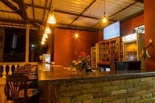 Uma área de bar com cadeiras de madeira, balcão de pedra, luminárias pendentes e uma geladeira.