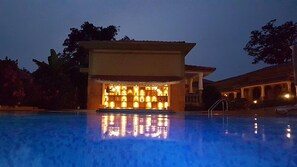 Um bar à beira da piscina com reflexo na água, um edifício com telhado em azulejos e céu claro.
