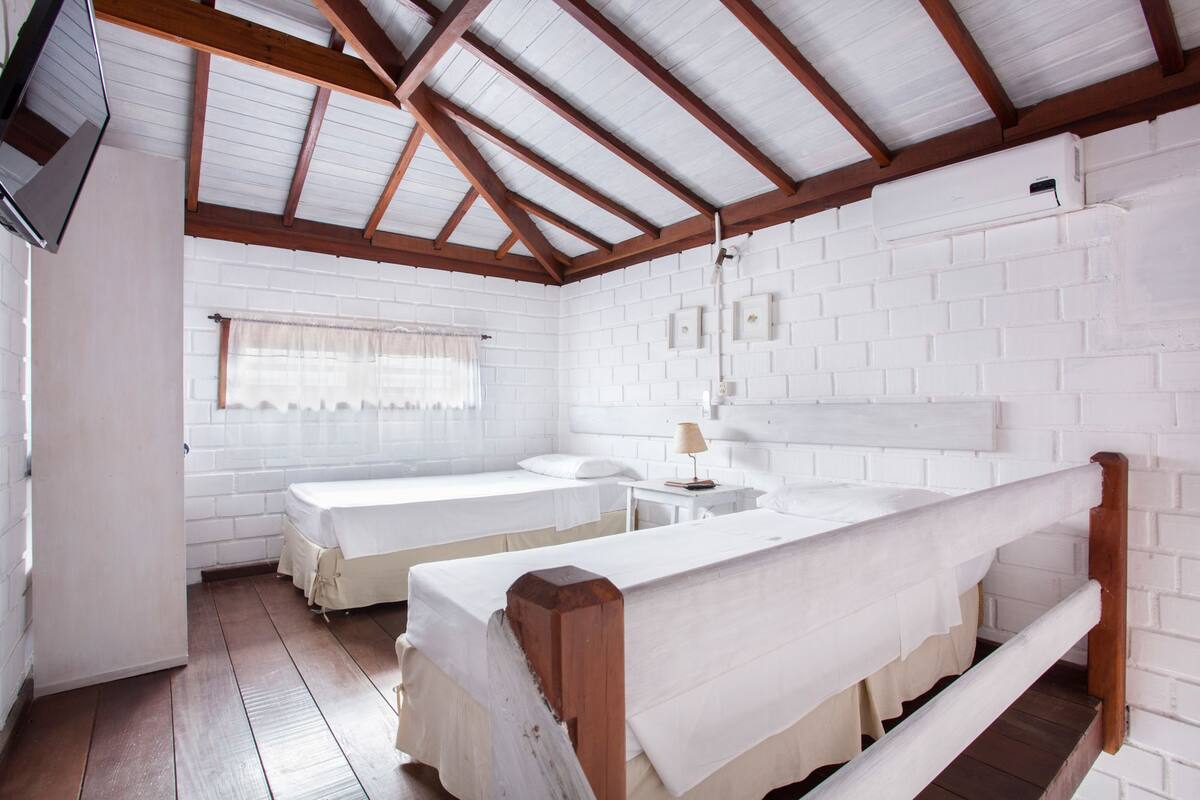 Quarto com duas camas de solteiro, teto de madeira, paredes de tijolo branco e janela com cortinas.