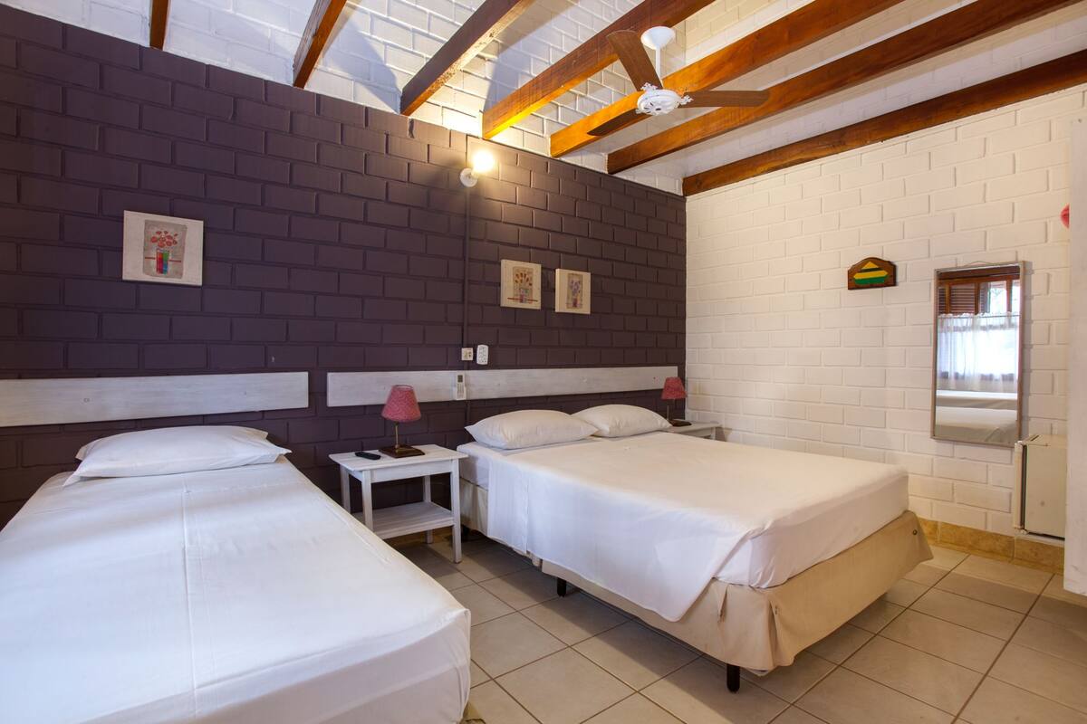 Quarto com duas camas, uma mesinha, um ventilador de teto e uma parede de tijolos brancos.