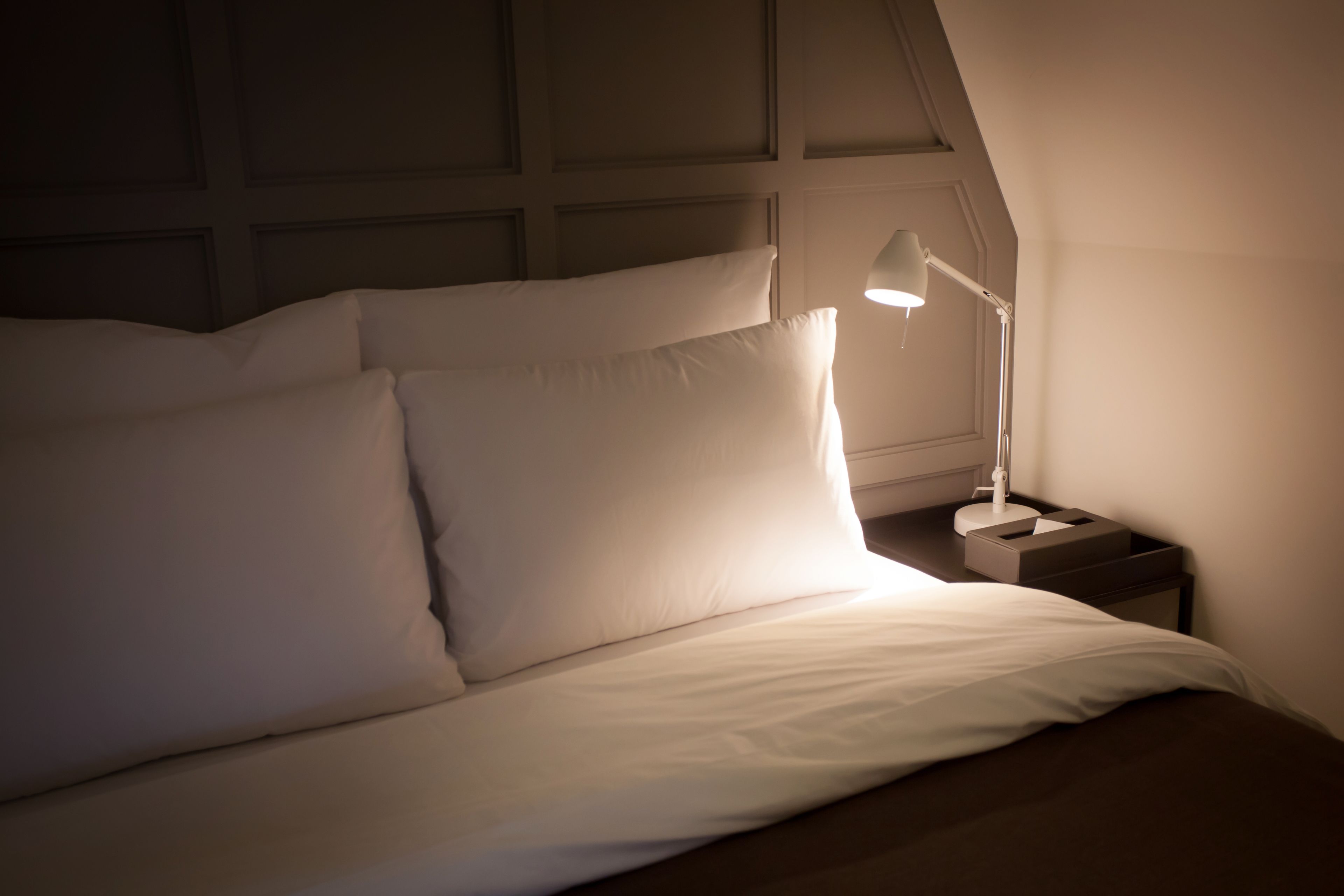 Cama bem arrumada com lençóis brancos em um quarto com pouca luz e uma luminária de cabeceira.