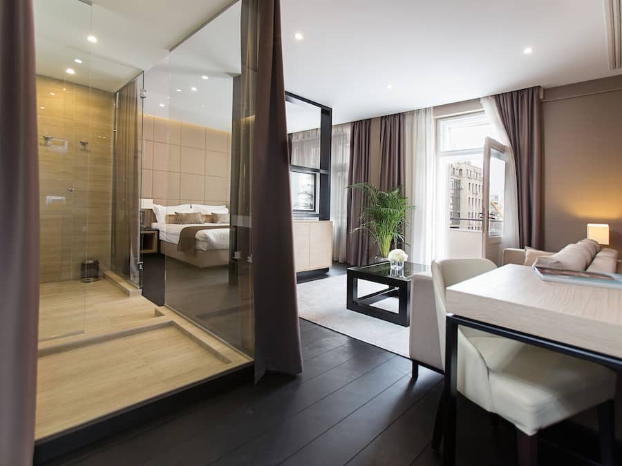 Quarto de hotel moderno com box de vidro, cama com roupa branca, área de refeições com mesa e cadeiras, e vista da cidade através de grandes janelas.