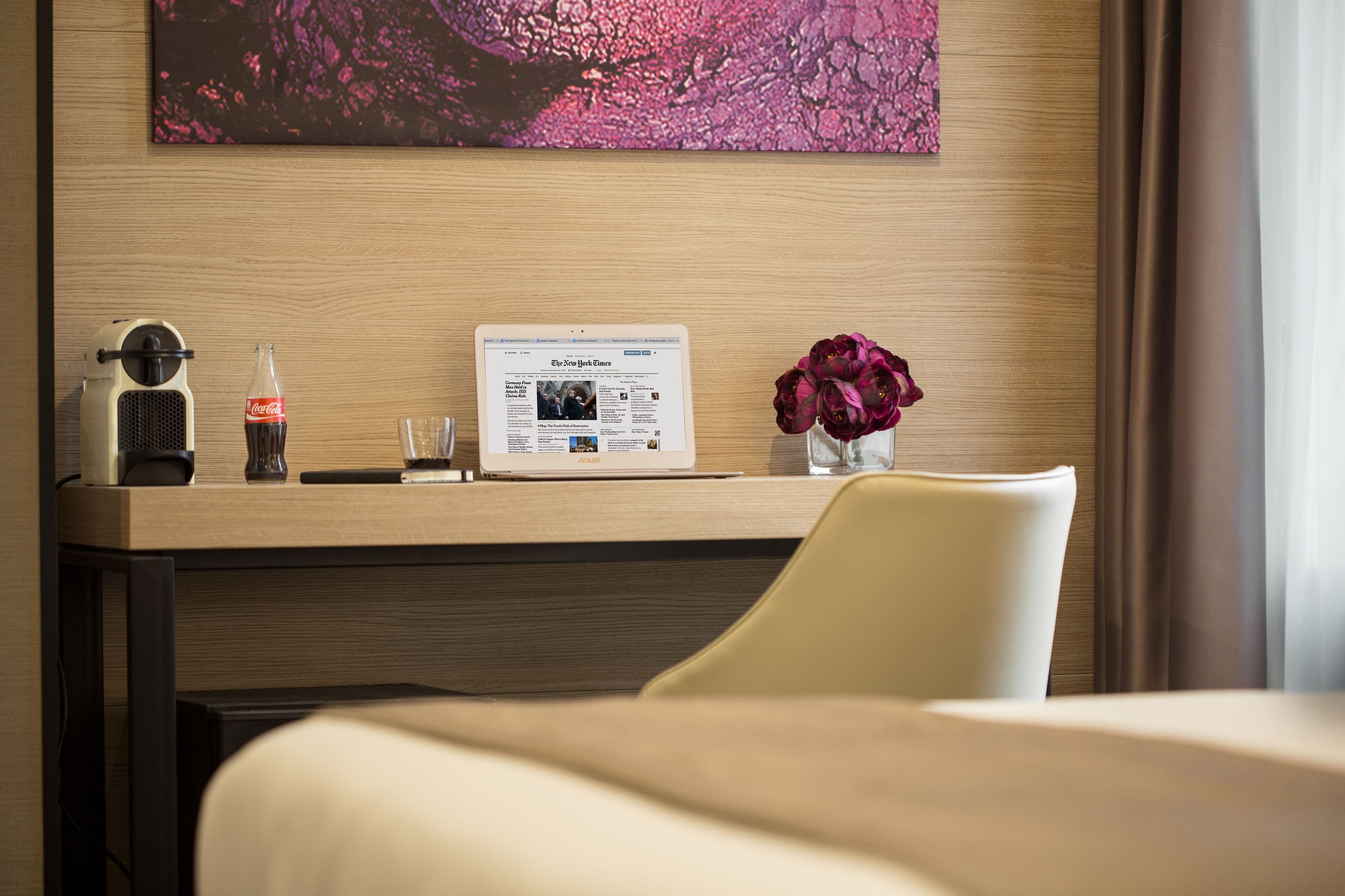 Quarto de hotel com uma secretária de madeira, jornal, garrafa de Coca-Cola, um vaso com flores e uma cadeira.