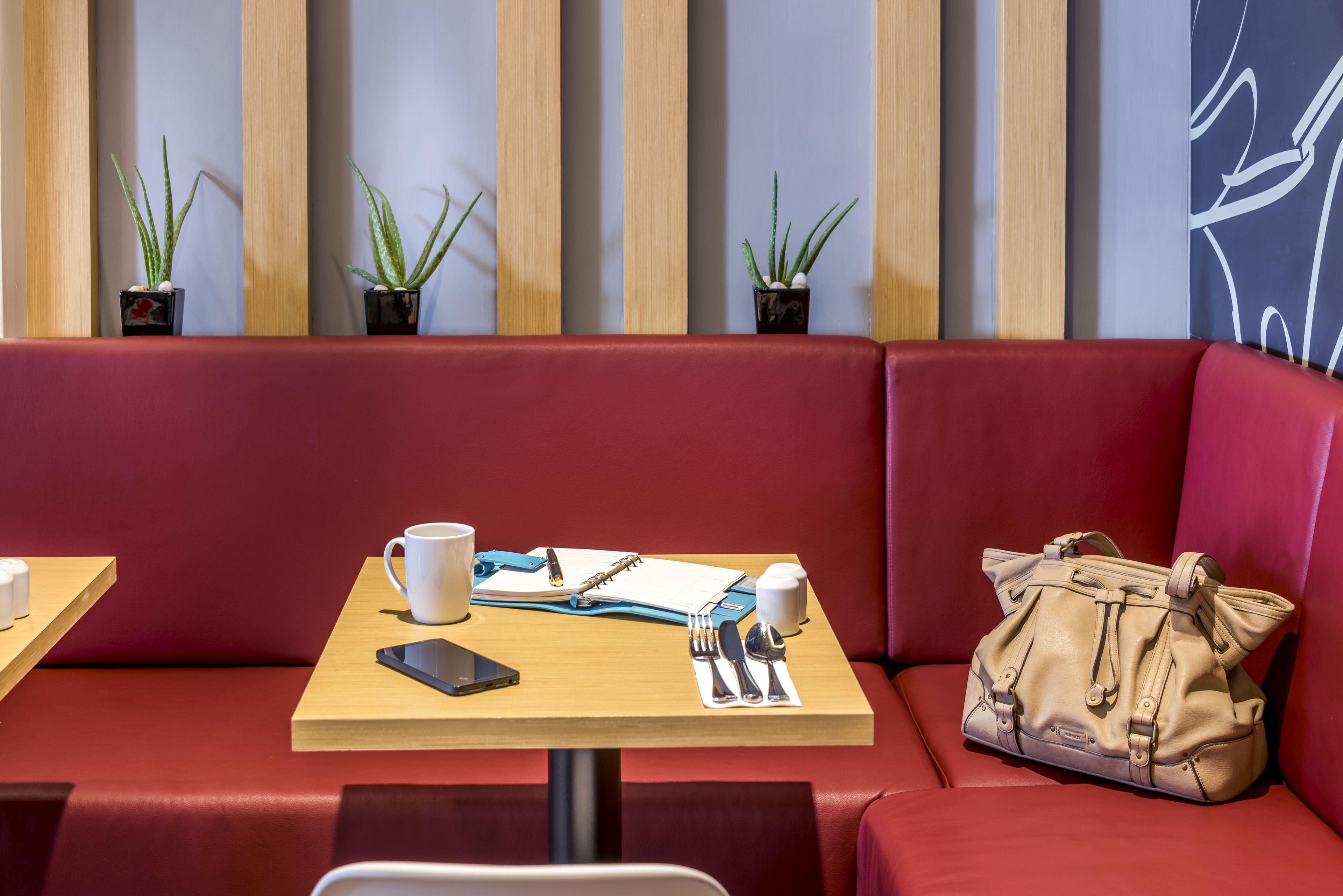 Um banco acolchoado vermelho com uma mesa de madeira na frente, uma bolsa bege e plantas em vasos na parede.