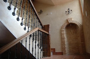 Uma escadaria com guarda-mão de madeira e balaústres pretos, conduzindo a um arco de pedra.