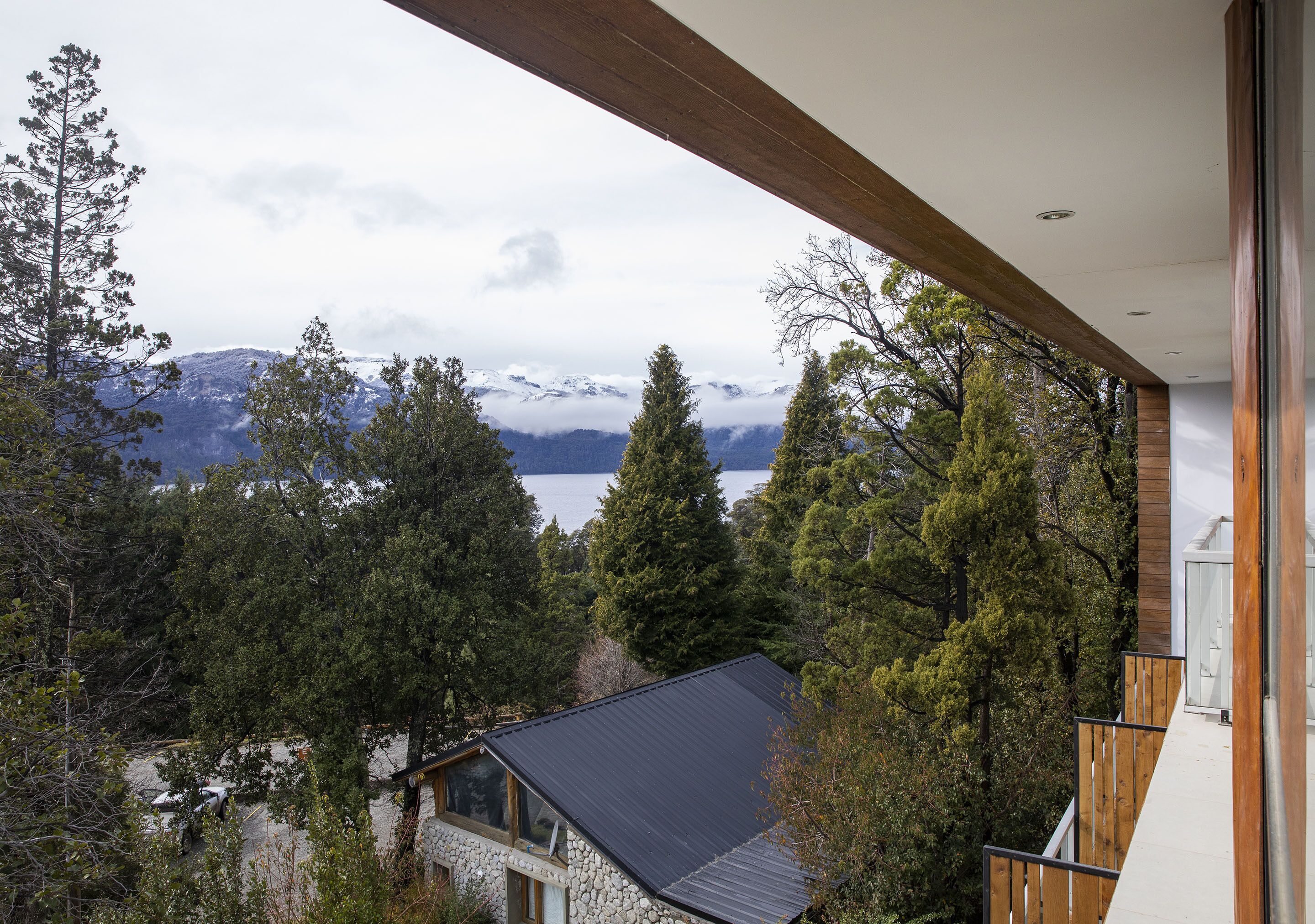 Uma casa moderna com fachada de pedra, telhado escuro e varanda com vista para uma área arborizada com um lago e montanhas ao fundo.