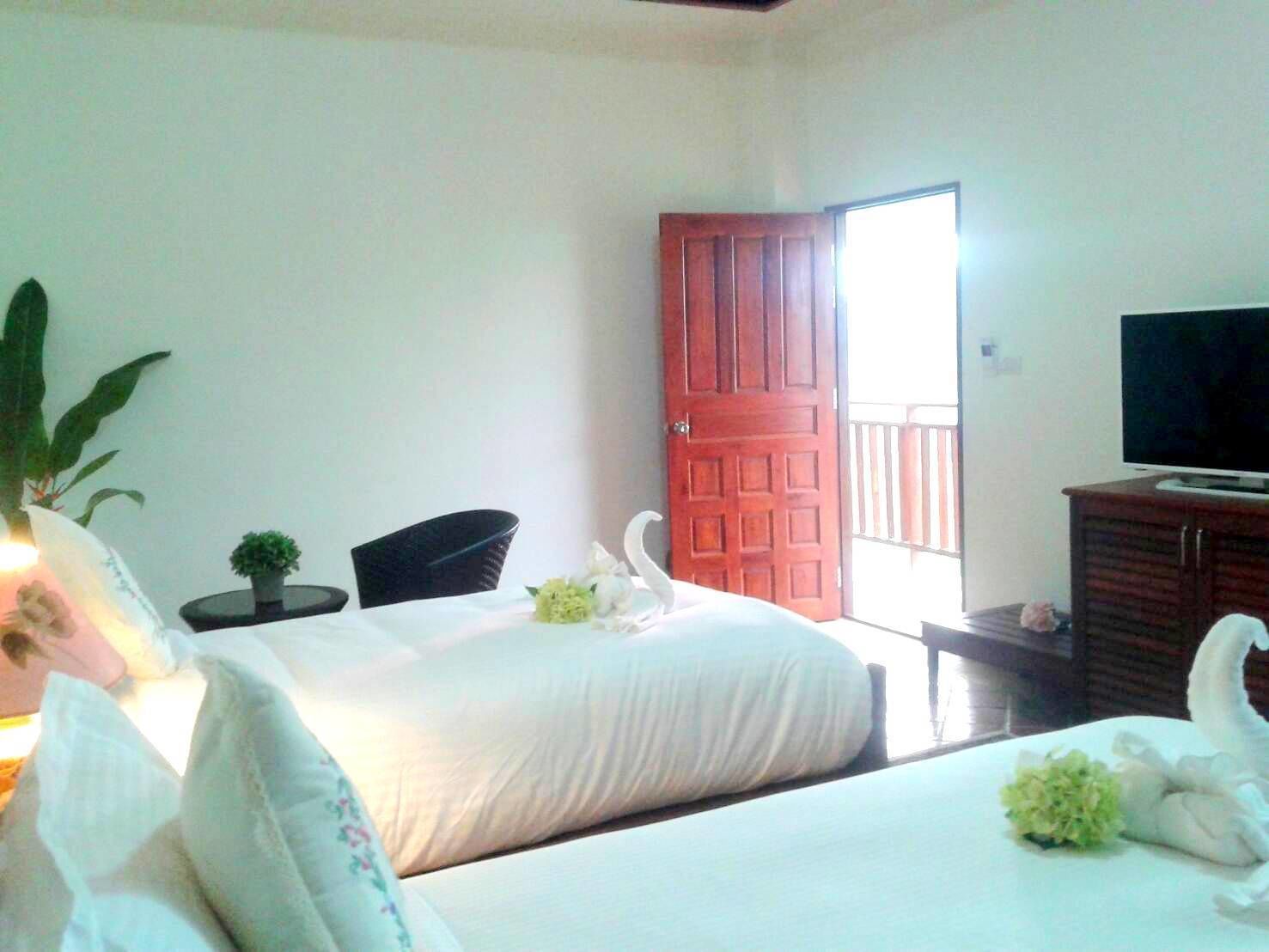 Quarto de hotel com duas camas, uma televisão, um armário de madeira, uma mesinha com uma planta e uma porta para o exterior.