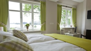 Quarto de hotel com uma cama grande, mesa de cabeceira com abajur, janela com cortinas e vista para o exterior.