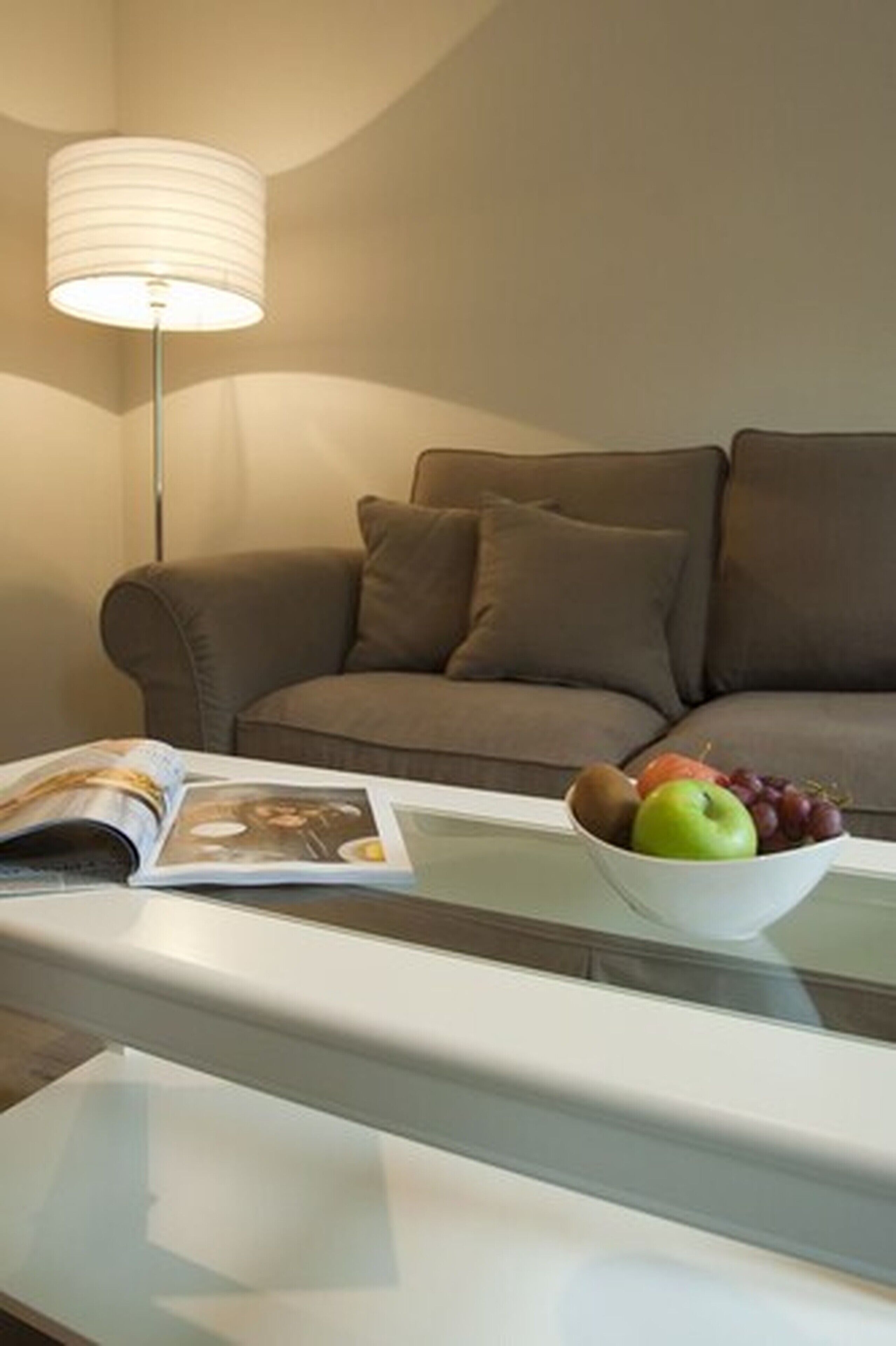 Uma sala de estar com sofá, abajur, revista e uma tigela de frutas sobre uma mesa de centro.