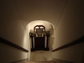 Um corredor com piso estampado que leva a uma porta de madeira escura, ladeada por duas colunas com arranjos florais.