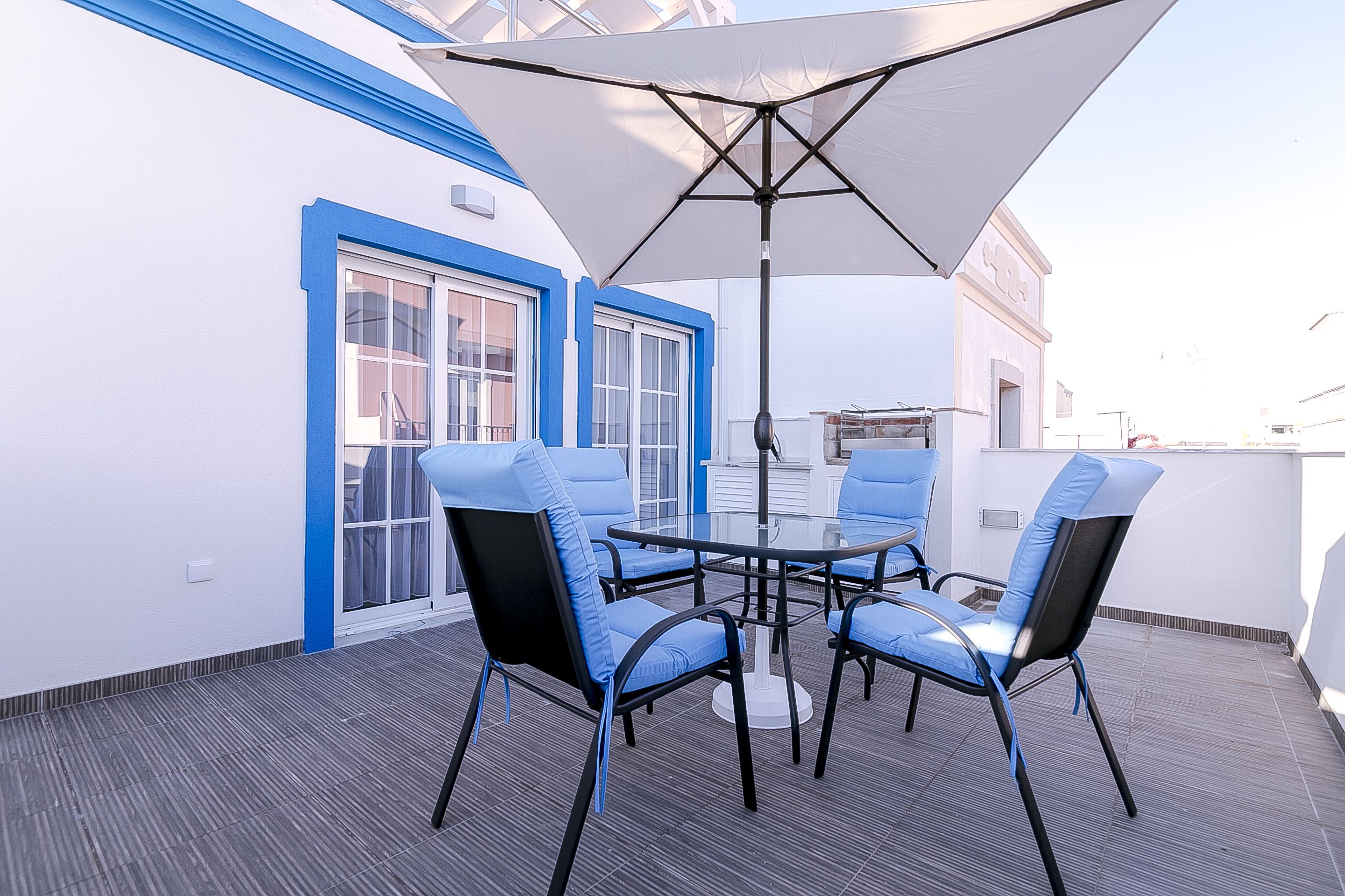 Um terraço com uma mesa redonda e quatro cadeiras, um guarda-sol e vista para a cidade.