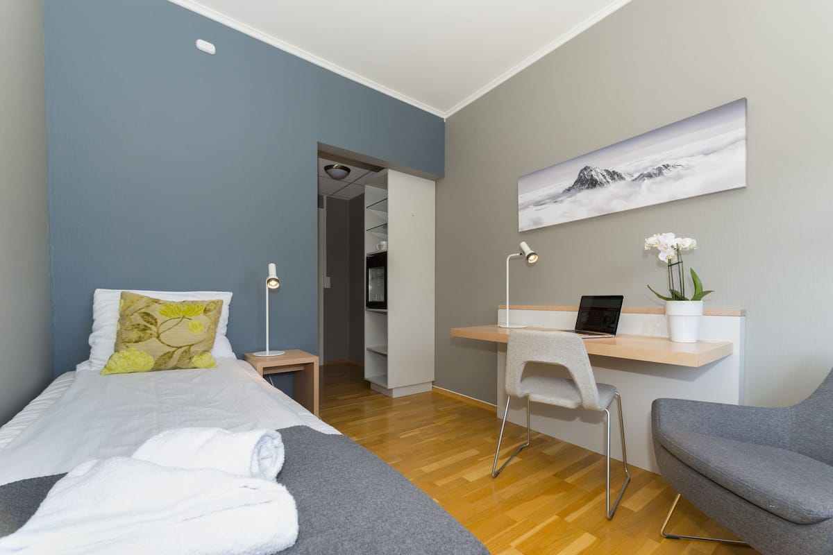 Quarto de hotel moderno com cama, escrivaninha, cadeira, luminária de mesa e planta em vaso.