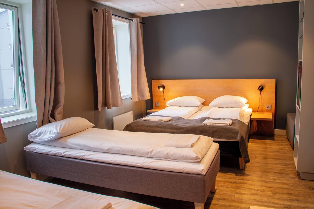 Quarto de hotel com duas camas, cabeceira de madeira, luminárias laterais e uma janela com cortinas.