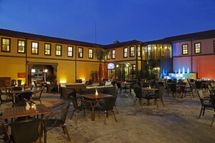 Uma área de café ao ar livre com mesas e cadeiras, um espaço de bar com balcão, e um edifício com várias janelas.
