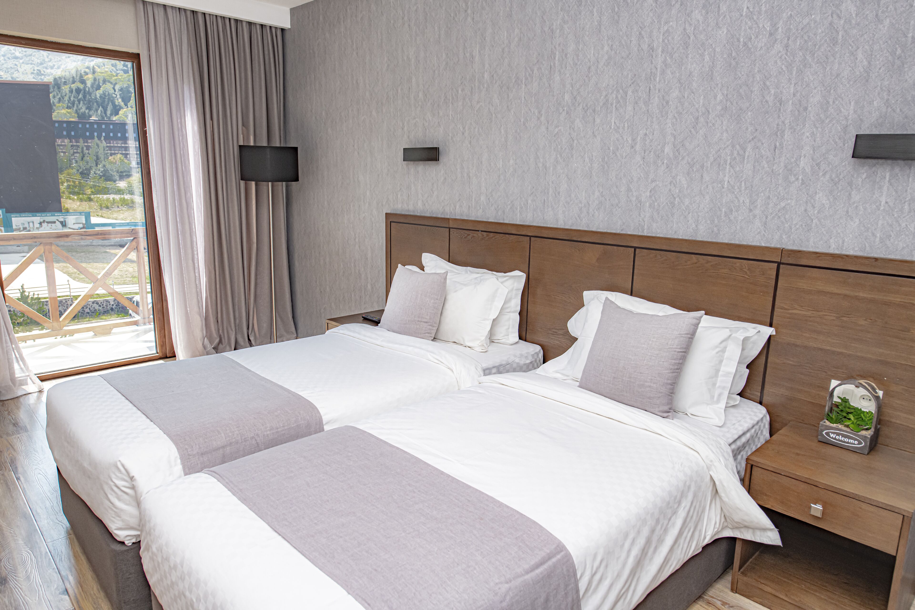 Quarto de hotel com duas camas, cabeceira de madeira, mesa de cabeceira com abajur e vista para o exterior.