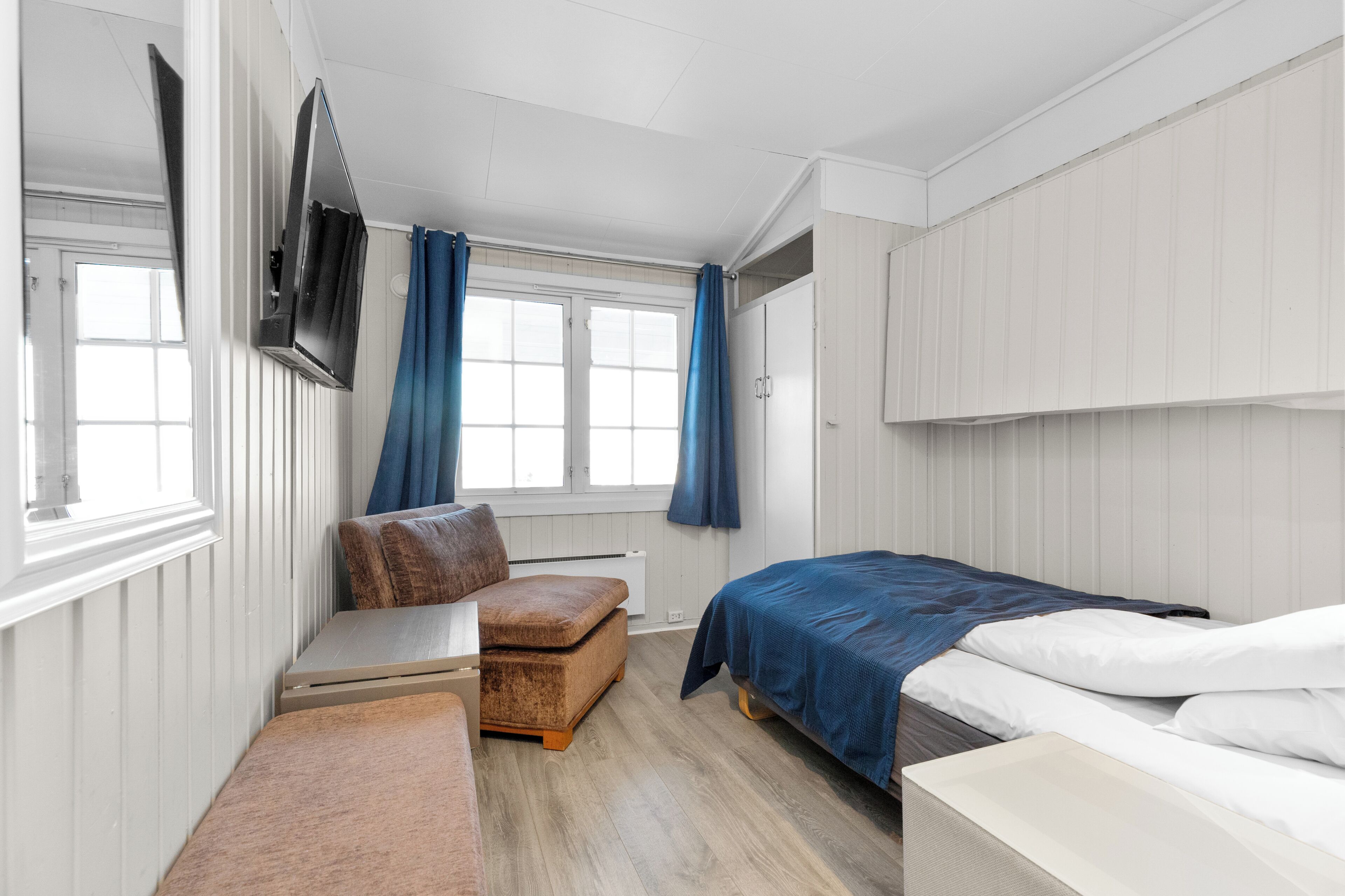 Quarto com cama, um banco, televisão fixada na parede e janela com cortinas.