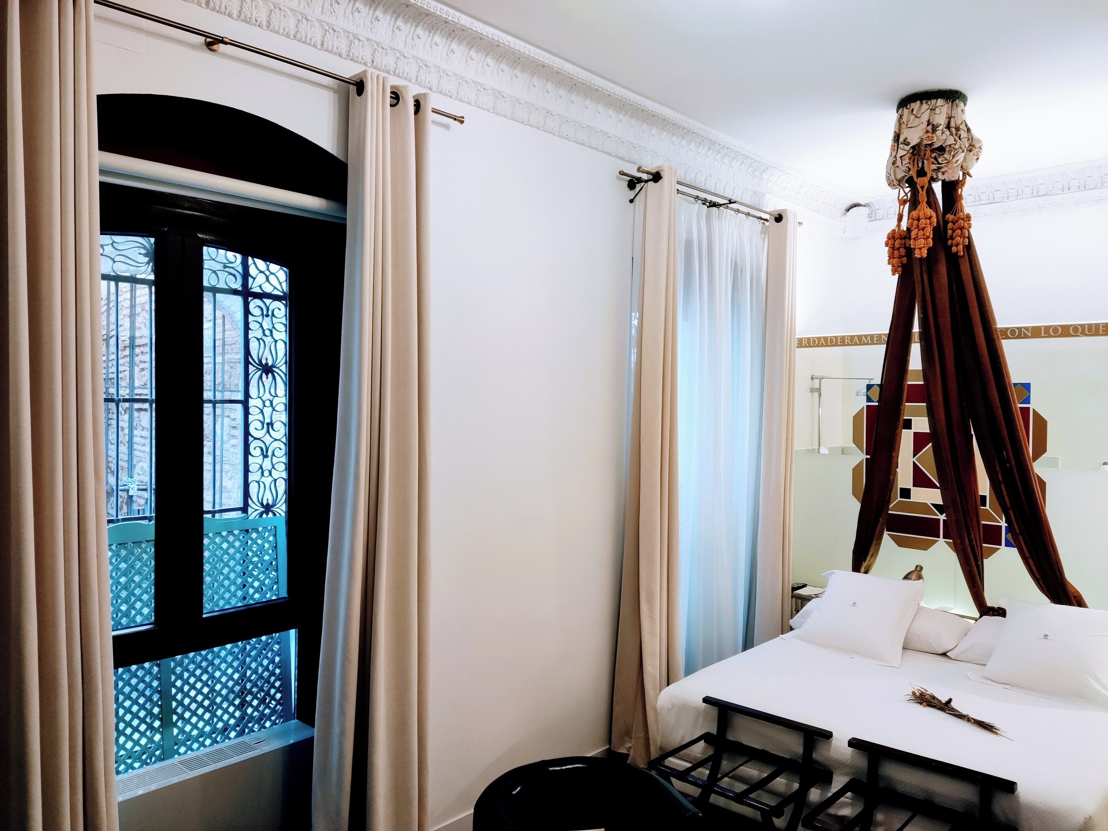 Quarto de hotel com cama, janela com cortinas, lustre e parede com padrão geométrico.