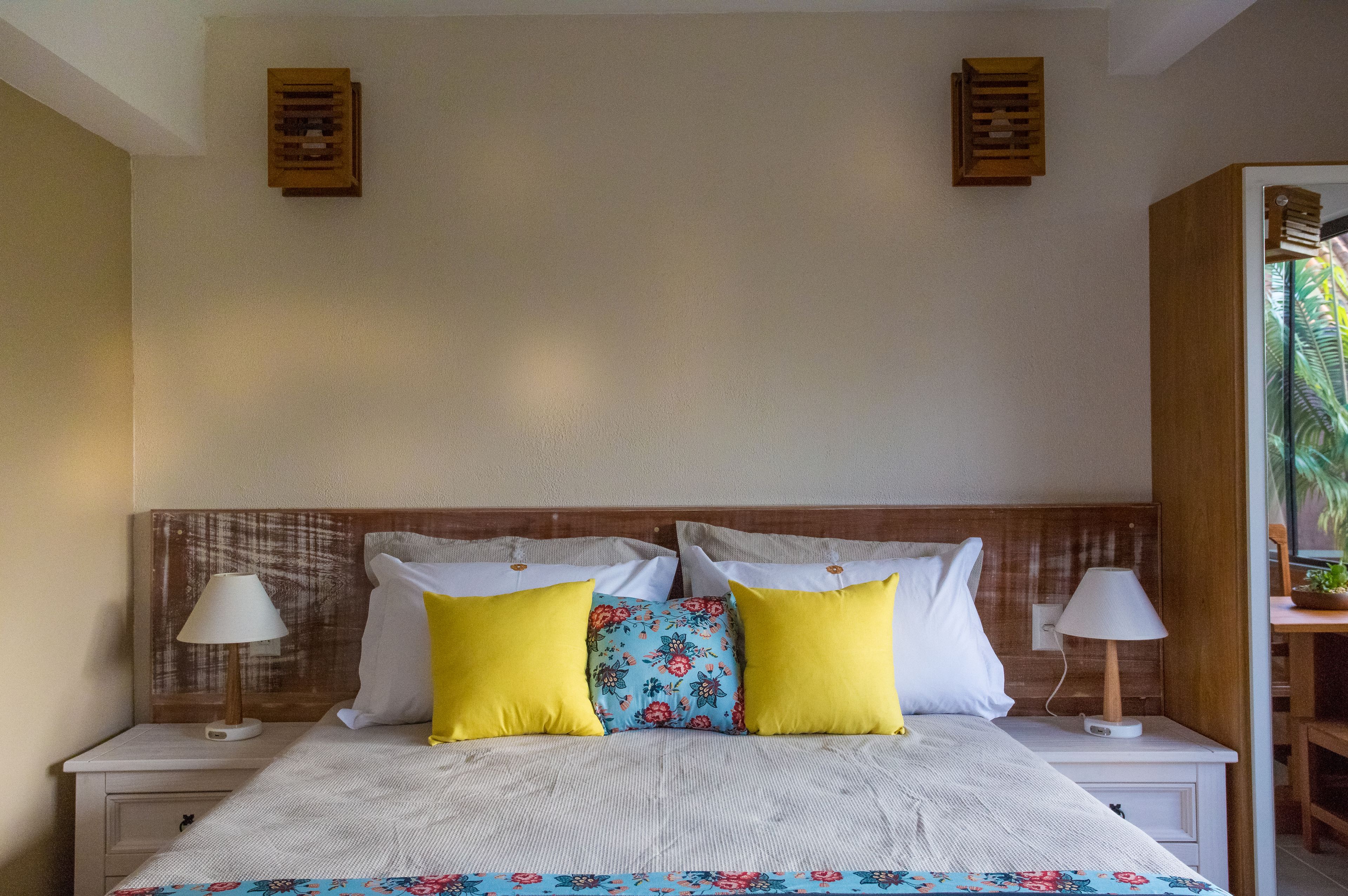 Cama bem arrumada com colcha estampada em flores, dois travesseiros brancos e dois amarelos. Há duas mesas de cabeceira com luminárias, um painel de madeira e uma pequena estante com uma planta.