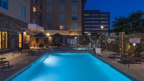 Piscina com espreguiçadeiras e guarda-sóis, cercada por edifícios de tijolo e uma área murada.