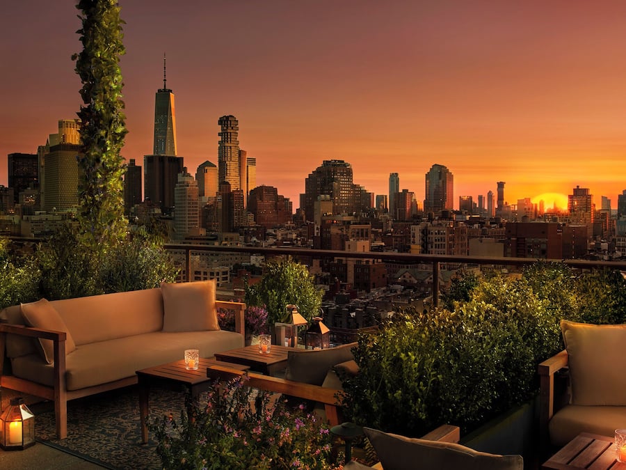 Terraço no telhado com vista para o horizonte da cidade ao pôr do sol, com um sofá, uma mesa de centro e plantas em vasos.