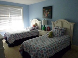 Quarto com duas camas, cada uma com roupa de cama e almofadas estampadas, uma mesa de cabeceira com abajur e relógio, e uma janela com persianas.