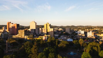 Paisagem urbana com diversos edifícios, incluindo um edifício vermelho em destaque e uma estrutura branca alta. Há uma área arborizada com árvores e uma fonte em primeiro plano.