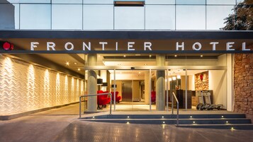 Exterior do Hotel Frontier com design moderno, apresentando entrada de vidro, fachada de pedra e uma área de estar vermelha visível no interior.