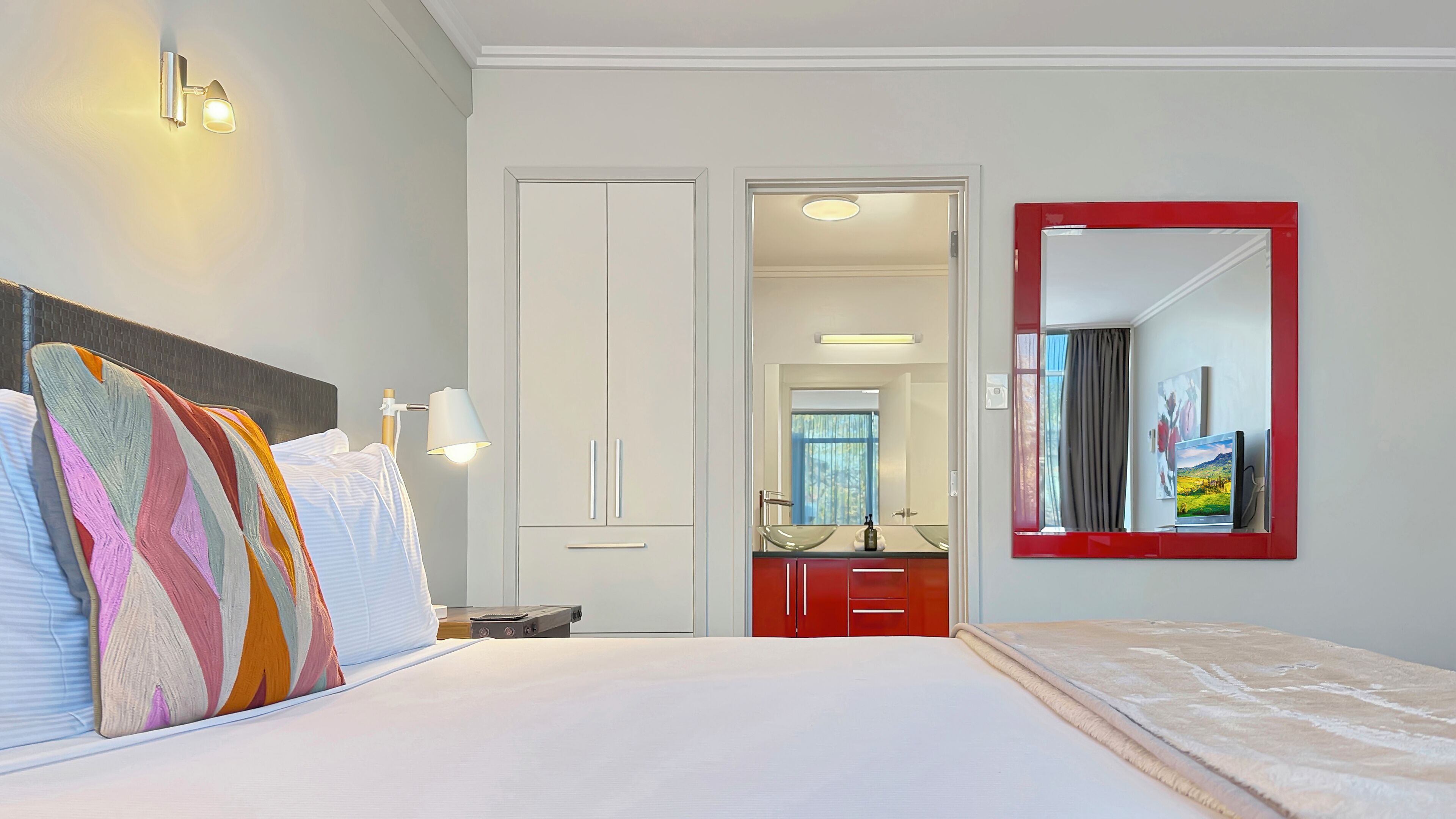 Quarto de hotel com uma cama grande, uma mesa de cabeceira com abajur, um espelho com moldura vermelha e vista para o banheiro.