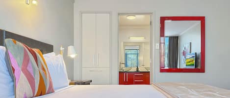 Quarto de hotel com uma cama grande, uma mesa de cabeceira com abajur, um espelho com moldura vermelha e vista para o banheiro.
