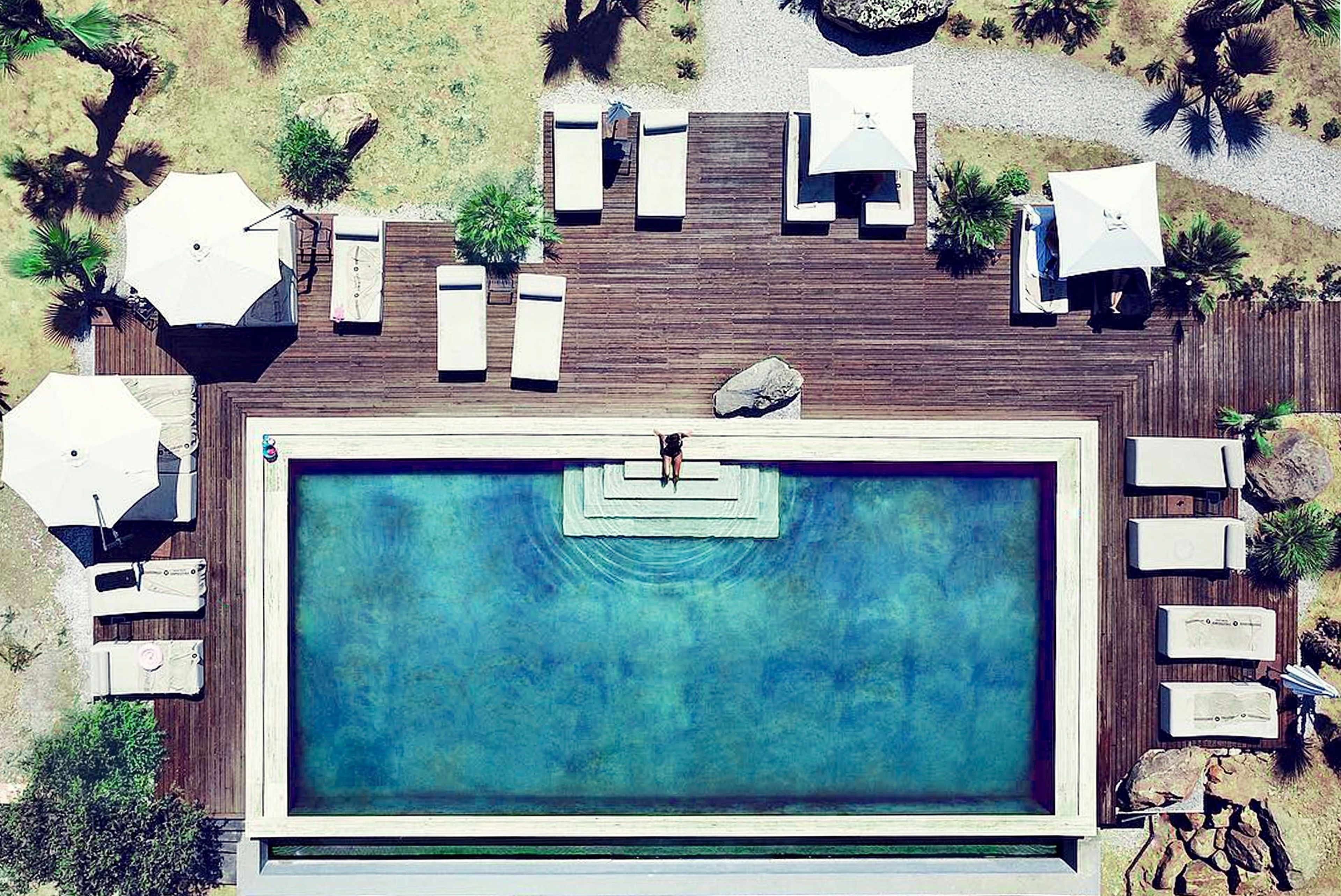 Vista aérea de uma piscina retangular com deck de madeira ao redor, equipada com espreguiçadeiras e guarda-sóis.