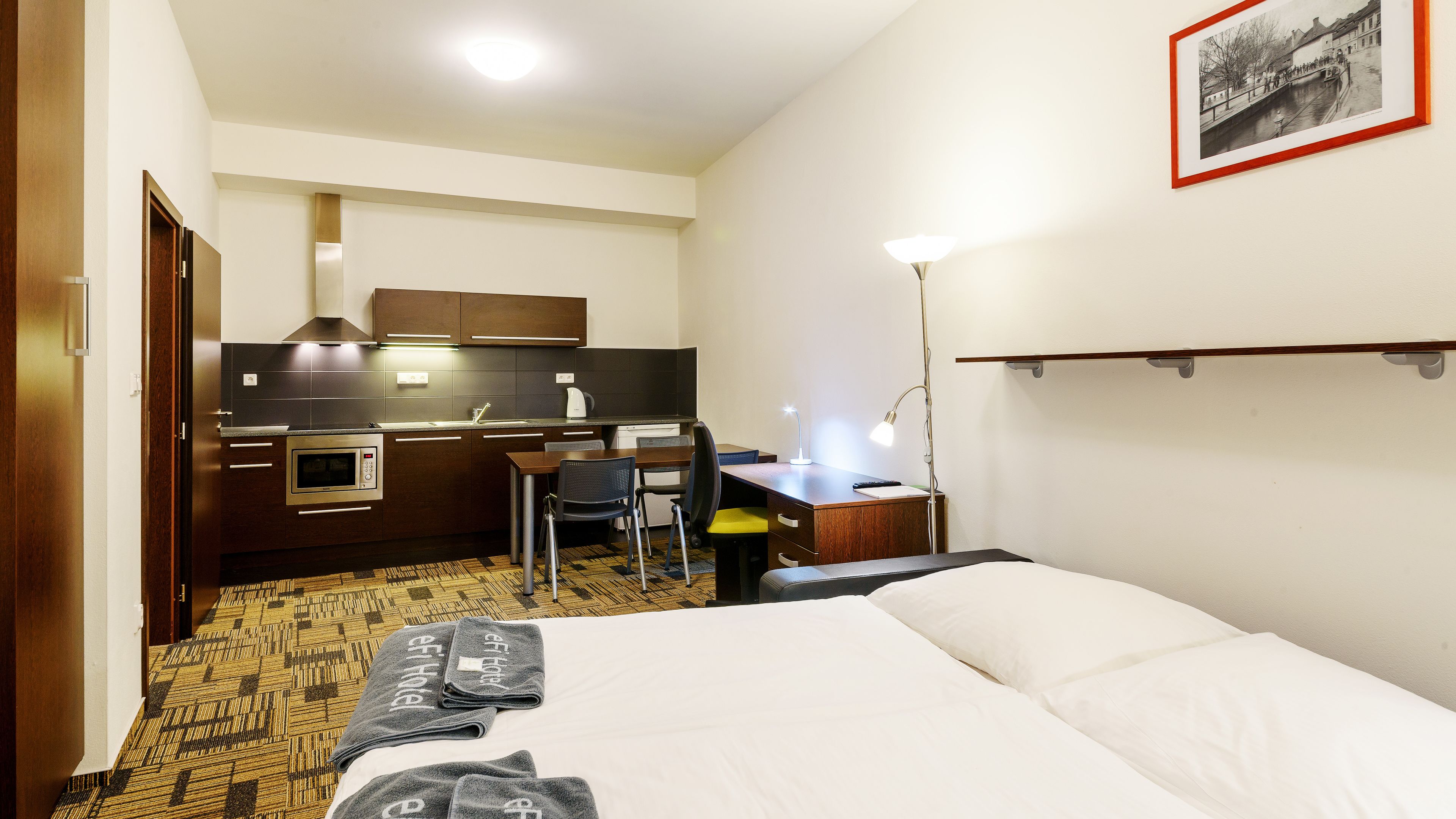 Quarto de hotel com cama, escrivaninha, cadeira, micro-ondas, pia e um quadro na parede.