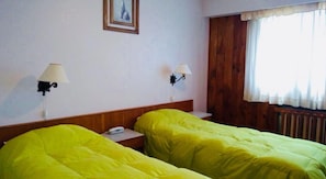 Quarto com duas camas, cada uma com um cobertor verde, uma cabeceira de madeira, uma mesa de cabeceira com abajur e uma janela com cortinas.