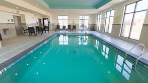 Uma piscina interior com água cristalina, rodeada por piso e paredes revestidas de azulejos, com janelas de um lado e uma área de estar com cadeiras do outro.