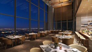 Restaurante moderno com pé-direito alto e grandes janelas, oferecendo vista para a cidade. Mesas dispostas com velas e arranjos de flores.
