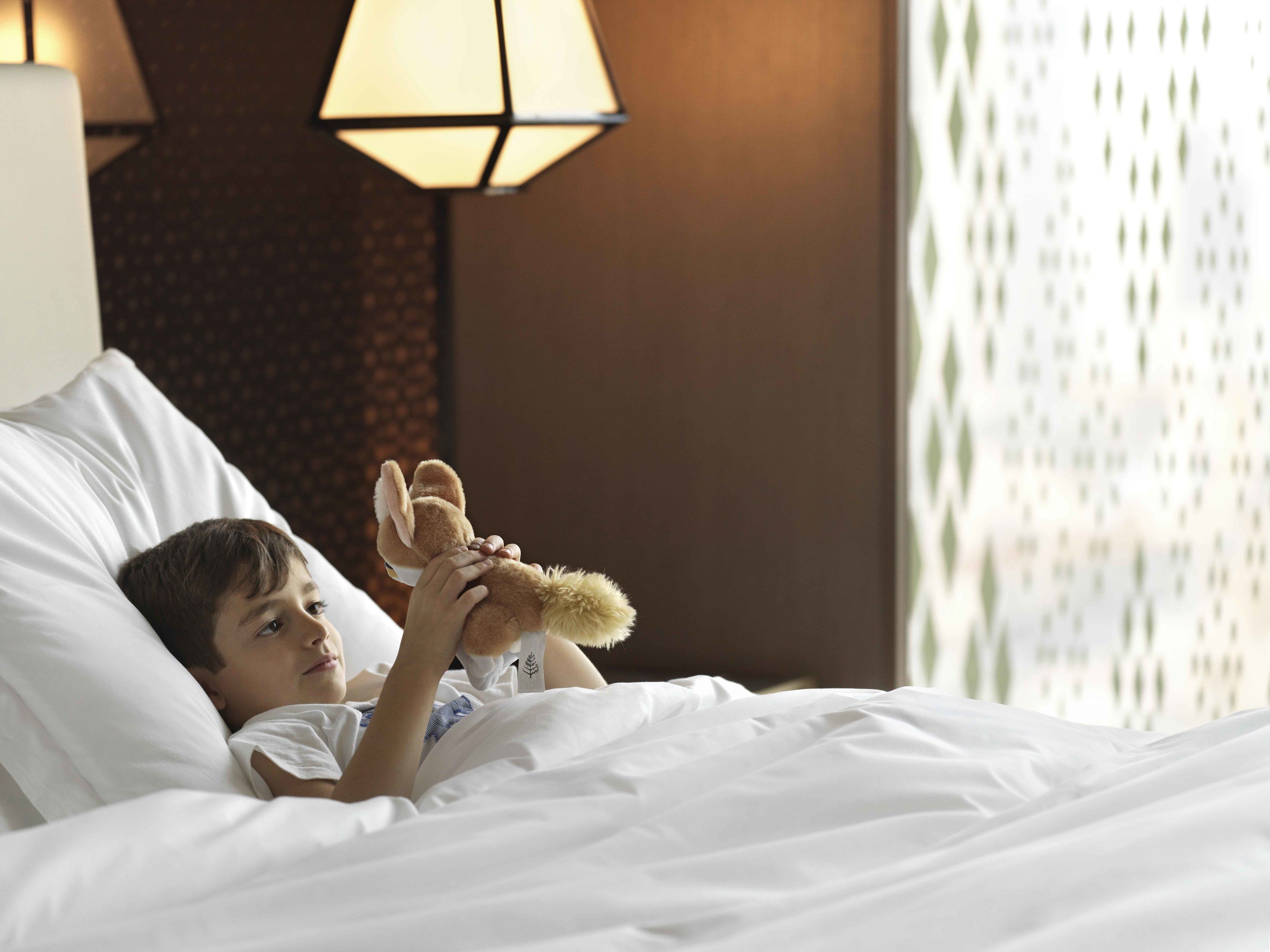 Criança deitada na cama com um coelho de pelúcia, roupa de cama branca e um abajur ao fundo.