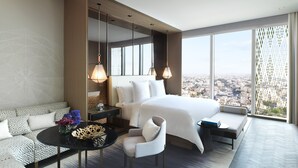Quarto de hotel moderno com uma cama grande, um sofá, uma pequena mesa e uma cadeira. Há uma janela ampla com vista para a cidade.
