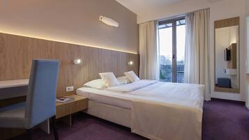 Quarto de hotel com uma cama, escrivaninha, cadeira, espelho e janela com cortinas.