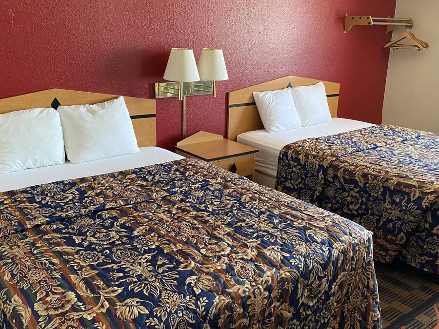 Quarto de hotel com duas camas, cada uma com colcha estampada de flores, uma mesa de cabeceira com abajur e uma parede vermelha.
