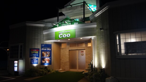 Um hotel simples chamado 'Coo', com uma placa verde, uma porta fechada e uma pequena área de jardim na frente.