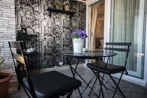 Uma pequena área externa com mesa redonda e duas cadeiras, um vaso de planta e uma parede decorativa com padrão de paisagem urbana.