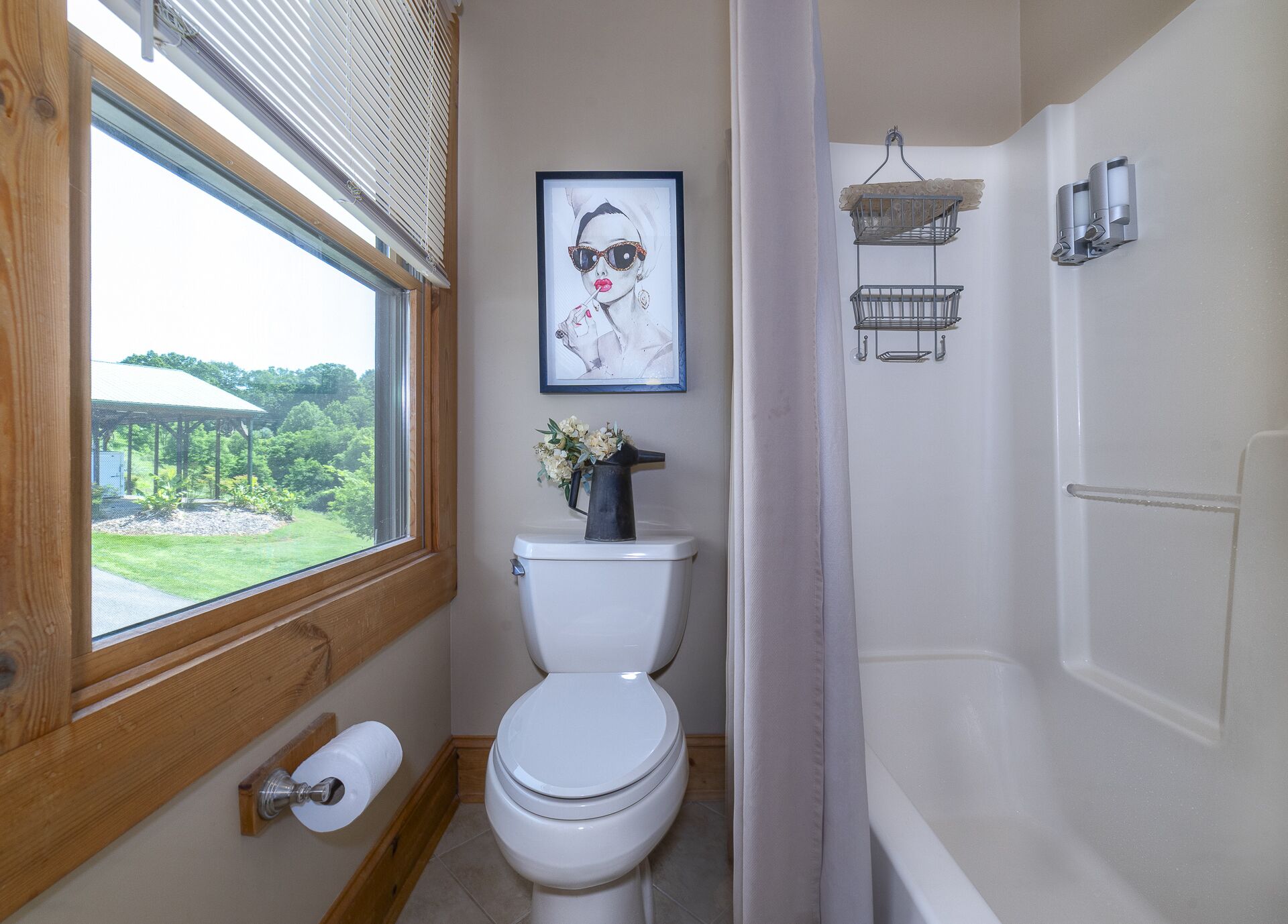Casa de banho com sanita, um quadro emoldurado acima, janela com vista para vegetação e zona de duche com porta-toalhas.