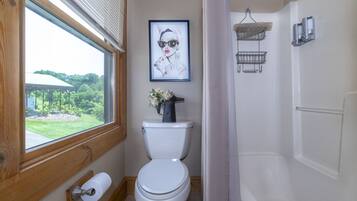 Casa de banho com sanita, um quadro emoldurado acima, janela com vista para vegetação e zona de duche com porta-toalhas.