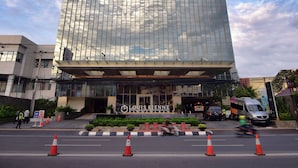 Um edifício alto e moderno com fachada de vidro refletiva, entrada imponente e céu claro com nuvens.
