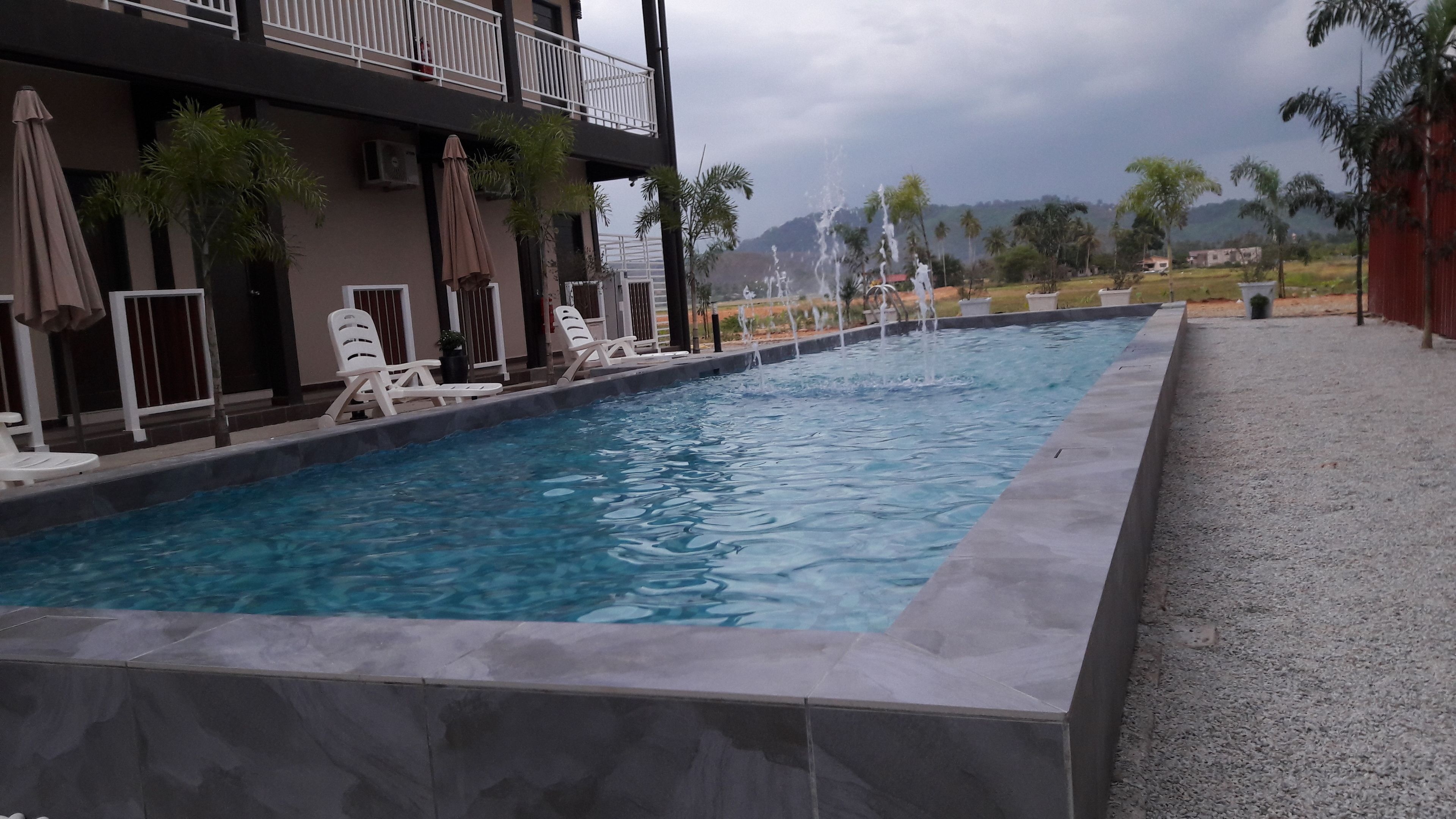 Uma piscina retangular com água azul cristalina, rodeada por um muro de pedra baixo. Há espreguiçadeiras e guarda-sóis brancos ao longo da borda da piscina.