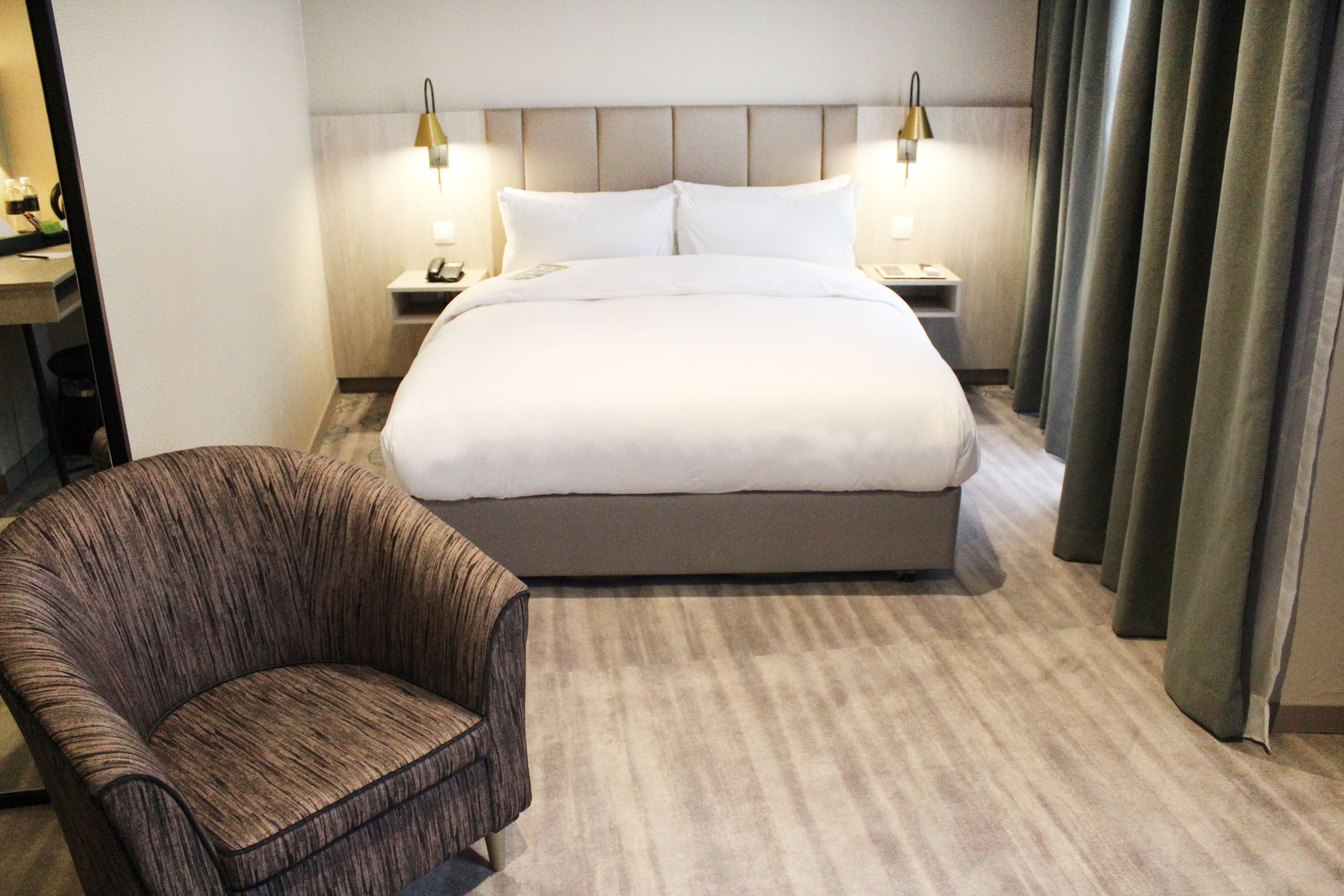 Quarto de hotel com uma cama grande, duas mesas de cabeceira com abajures, uma cadeira e uma cortina.